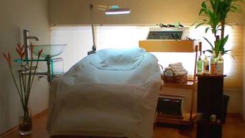 Quarto com cama coberta por lençol branco, escrivaninha com computador, luminária, planta e prateleira com artigos de higiene pessoal.