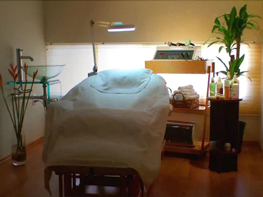 Quarto com cama coberta por lençol branco, escrivaninha com computador, luminária, planta e prateleira com artigos de higiene pessoal.