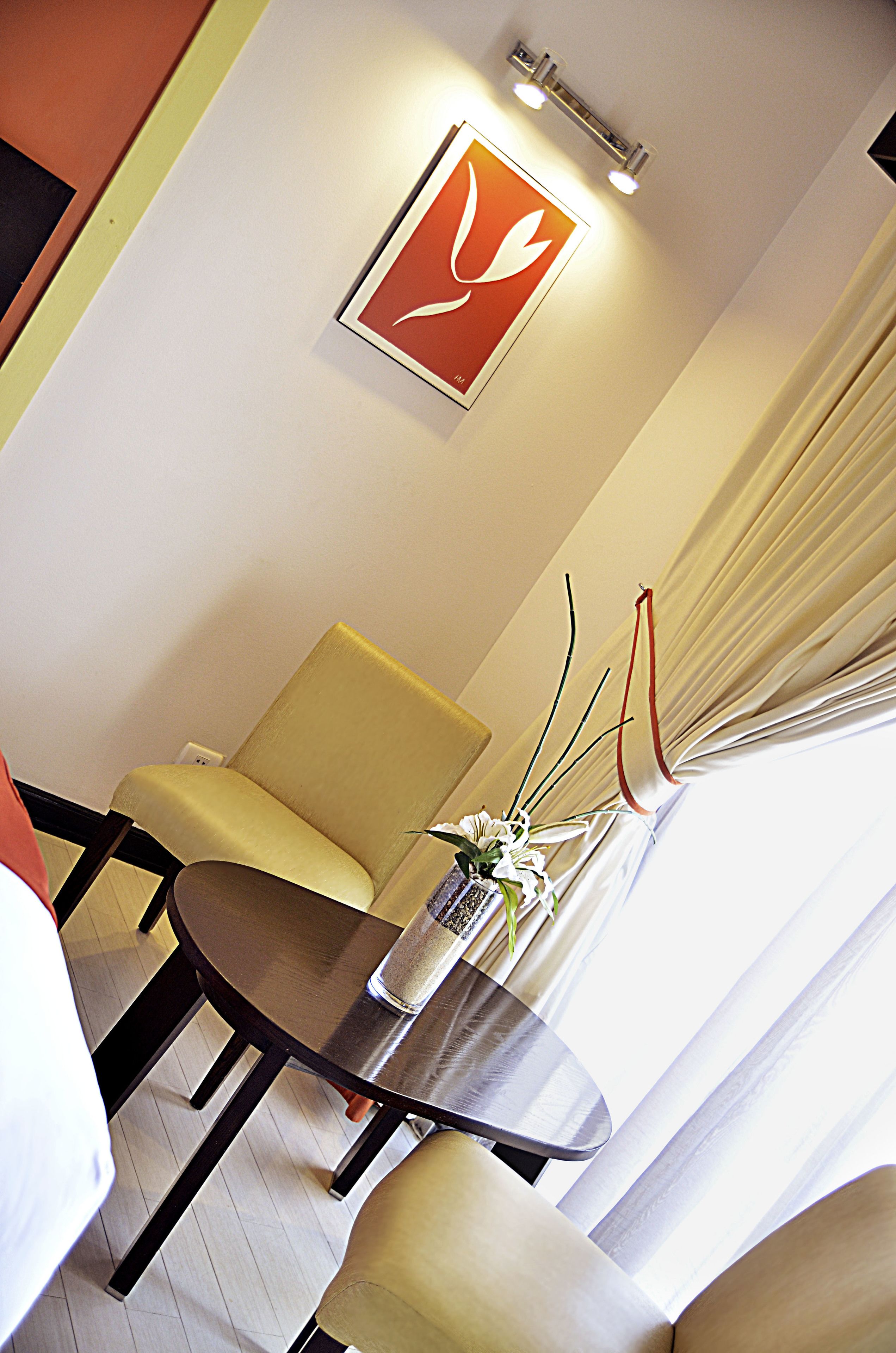 Quarto de hotel com uma mesa redonda, uma poltrona amarela, um vaso com flores e uma obra de arte emoldurada na parede.