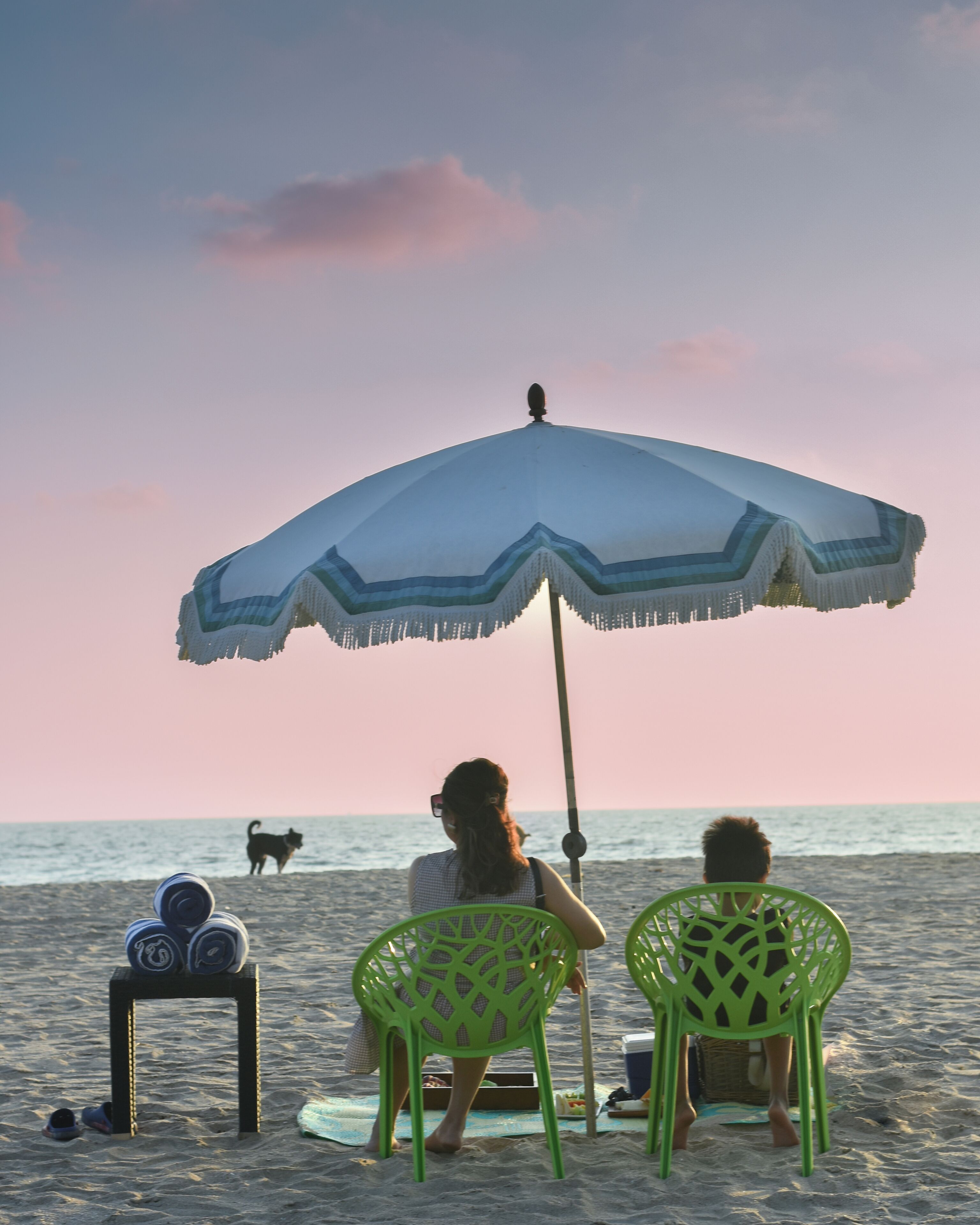 Cena de praia com duas pessoas sob um guarda-sol grande, um cão ao fundo e uma cadeira com objetos na areia.