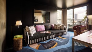 Quarto de hotel moderno com um sofá escuro, uma mesinha, uma luminária, um espelho e vista da cidade por grandes janelas.