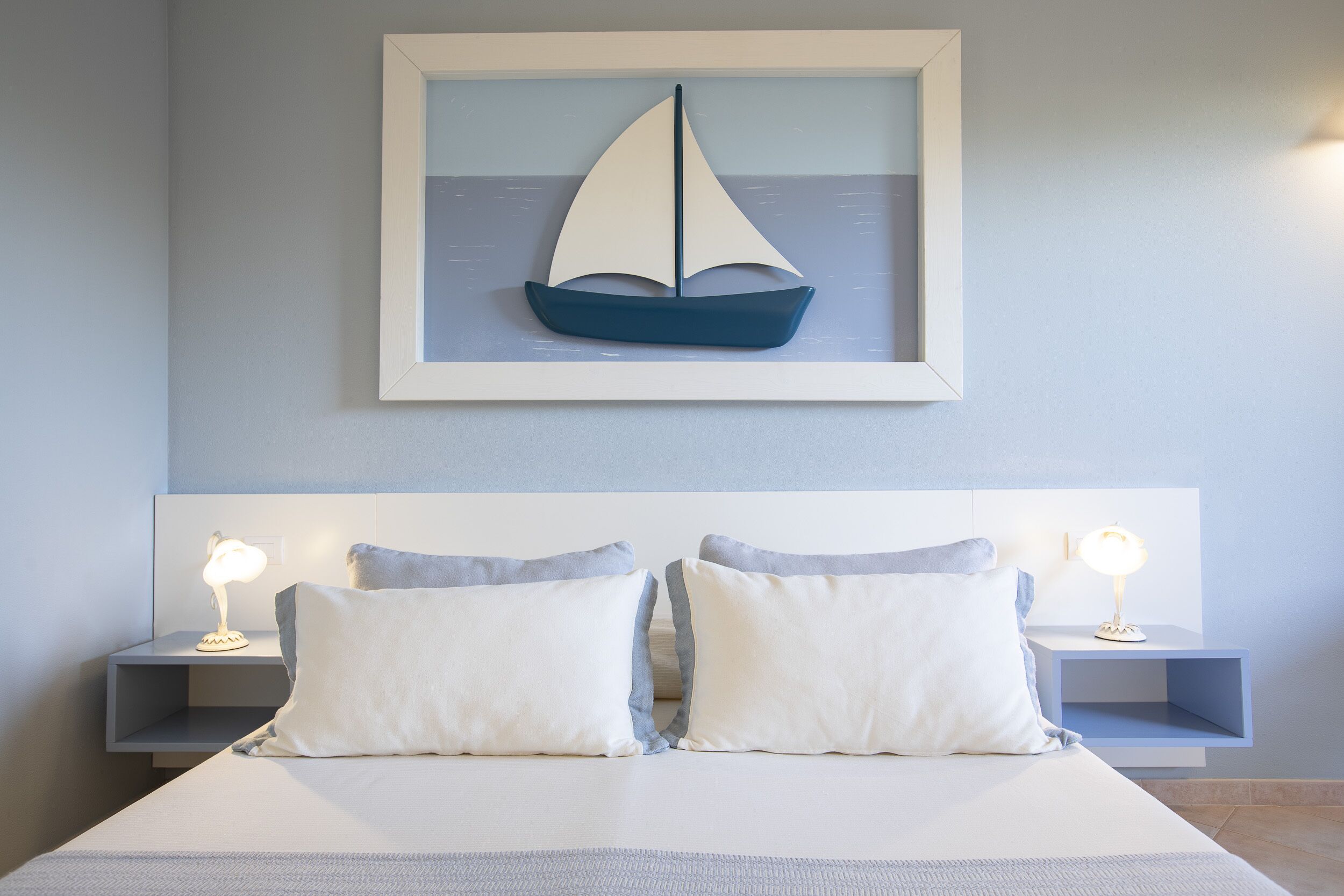 Quarto com cama, duas mesas de cabeceira com luminárias e um quadro de um veleiro na parede.