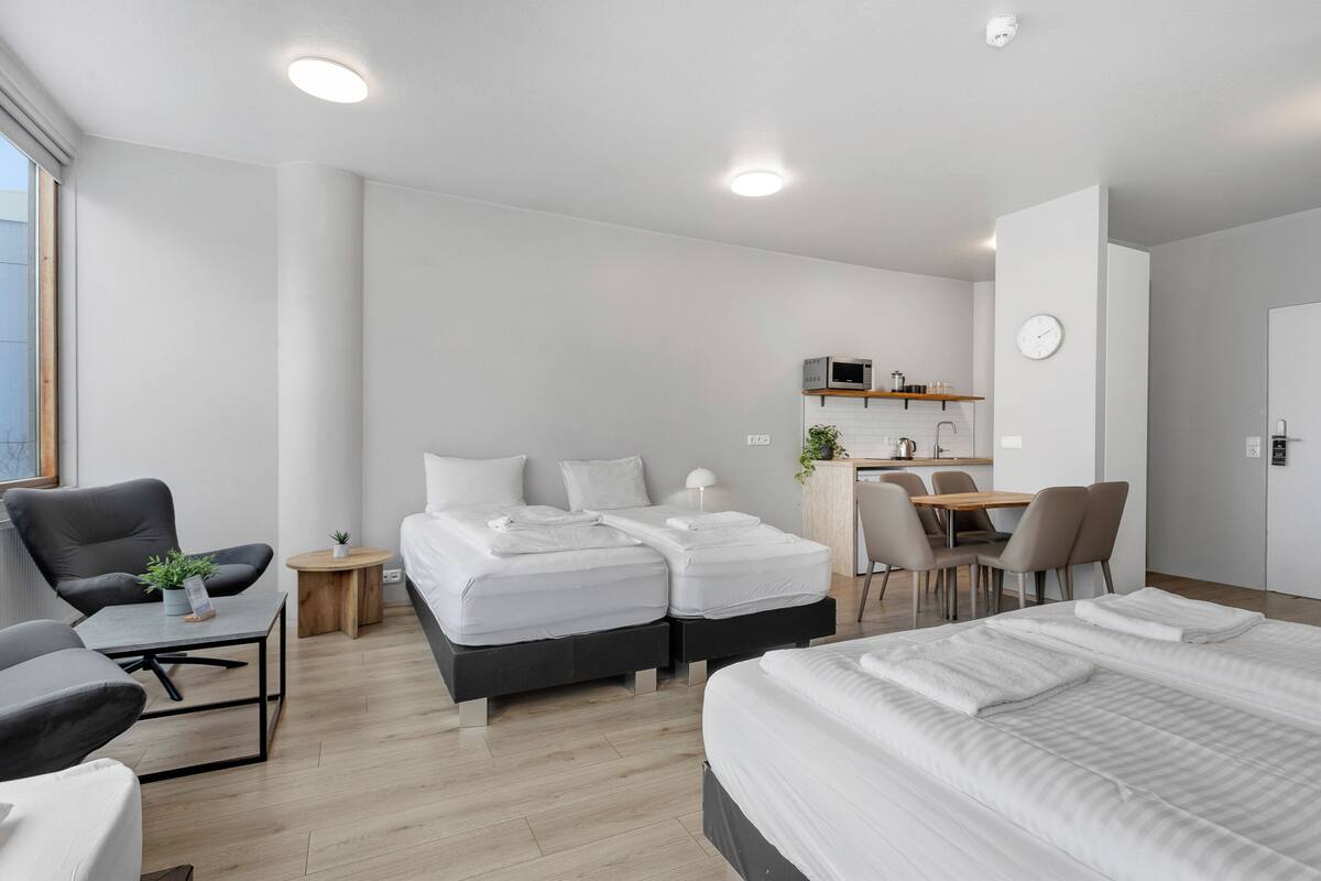 Quarto de hotel moderno com duas camas, uma escrivaninha, uma cadeira, uma mesinha com uma planta e uma janela.