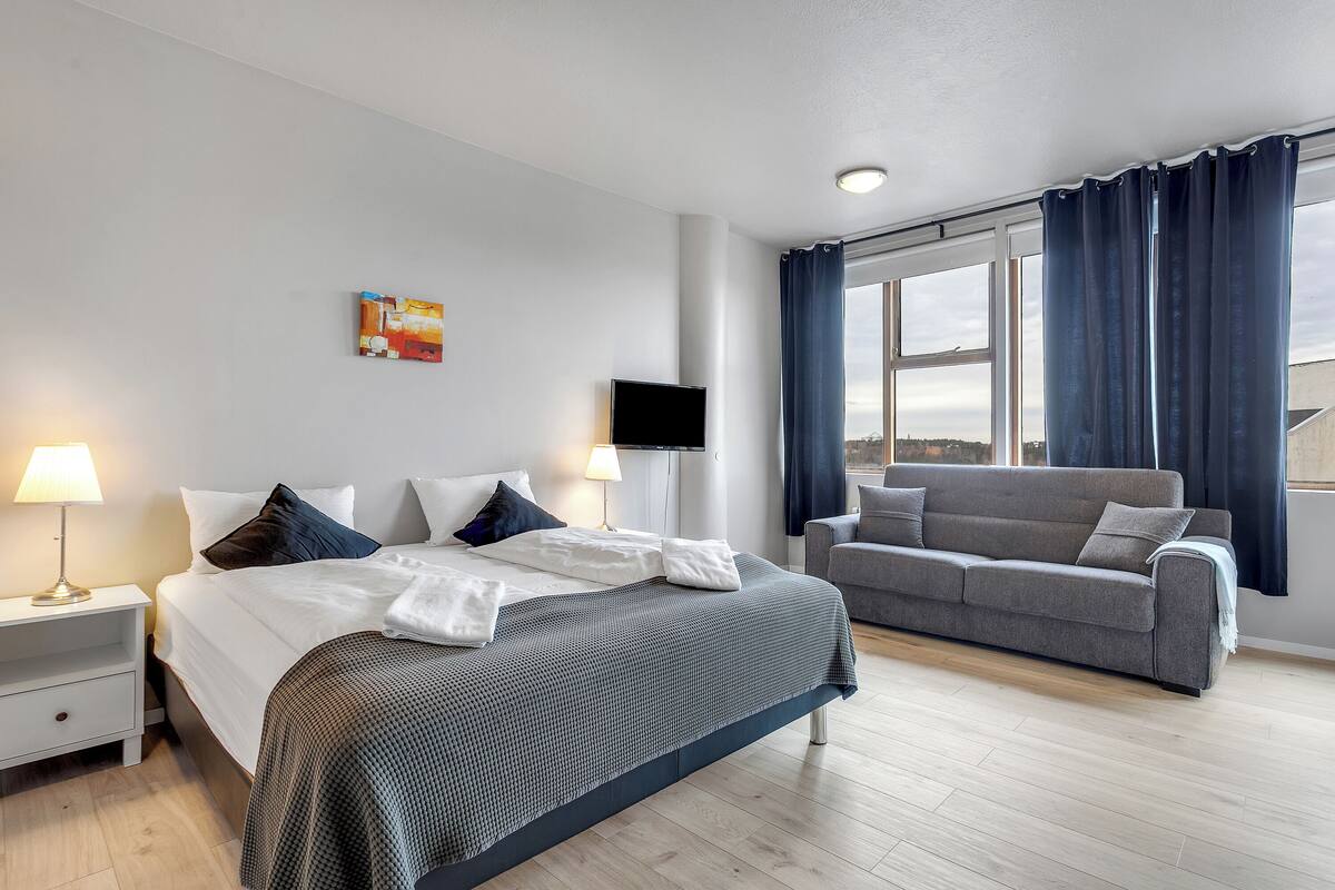 Quarto de hotel moderno com cama, sofá, televisão de tela plana e janela com cortinas.