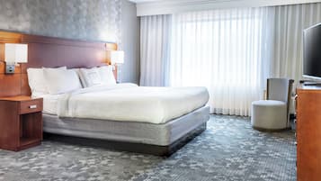 Quarto de hotel com uma cama grande, cabeceira de madeira, mesa de cabeceira, escrivaninha com cadeira, televisão e uma grande janela com cortinas translúcidas.