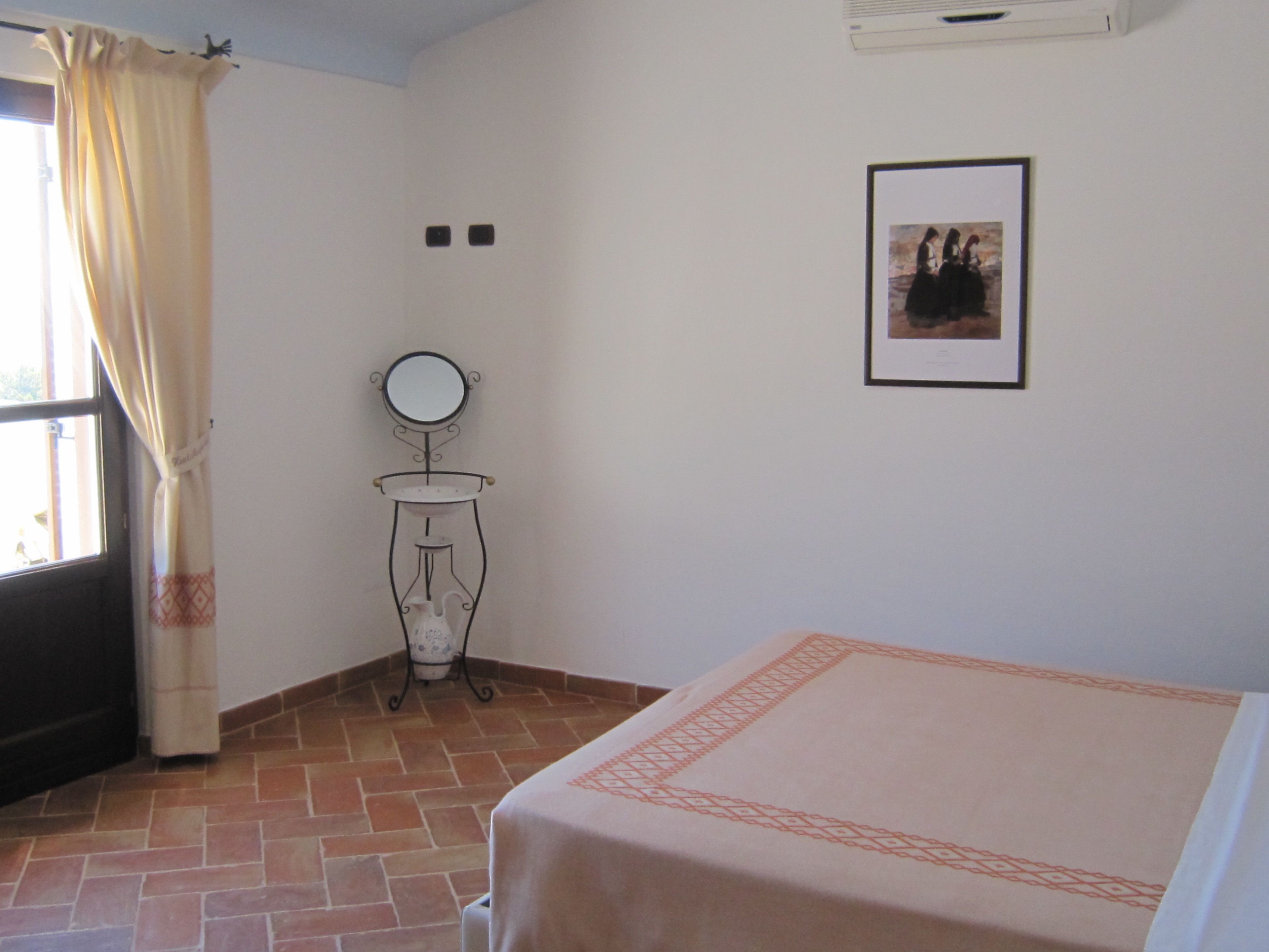 Quarto com cama, uma pequena mesa com espelho, um quadro na parede e uma janela com cortinas.