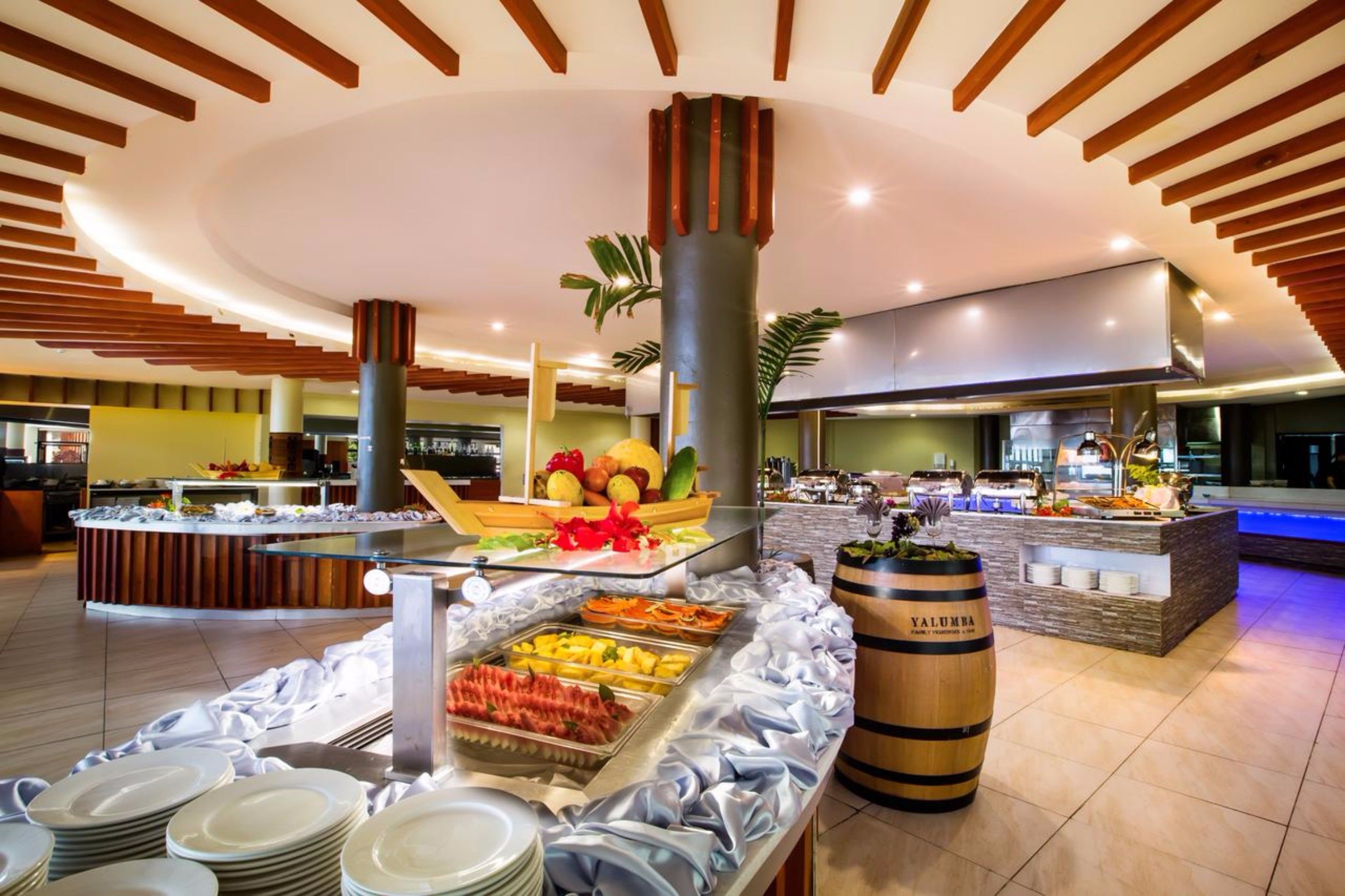 Uma área de buffet com diversos pontos de comida, um pilar decorativo central e um bar com um barril identificado como "VALUMBA".