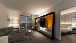 Uma sala de estar moderna com um sofá, uma mesa de centro e uma TV. Há uma janela grande com vista para a cidade.