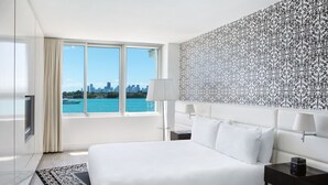 Quarto de hotel moderno com uma cama grande, vista para a cidade, parede com padrão e mesa de cabeceira com abajur.
