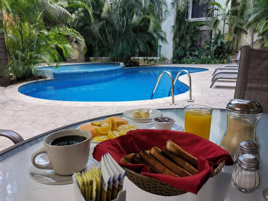 Mesa de café da manhã com café, fatias de laranja, pão e um copo de suco de laranja à beira da piscina.