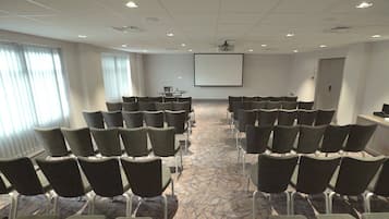 Uma sala de conferências com fileiras de cadeiras voltadas para uma tela de projeção, janelas amplas com persianas e piso coberto de carpete.
