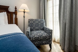 Quarto de hotel com uma cama, uma poltrona estampada, uma luminária de chão e uma janela com cortinas.