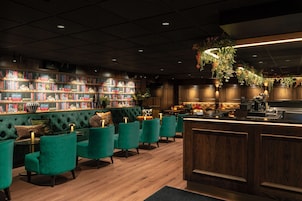 Uma área de bar com balcão de madeira, cadeiras estofadas verdes e uma estante cheia de livros.