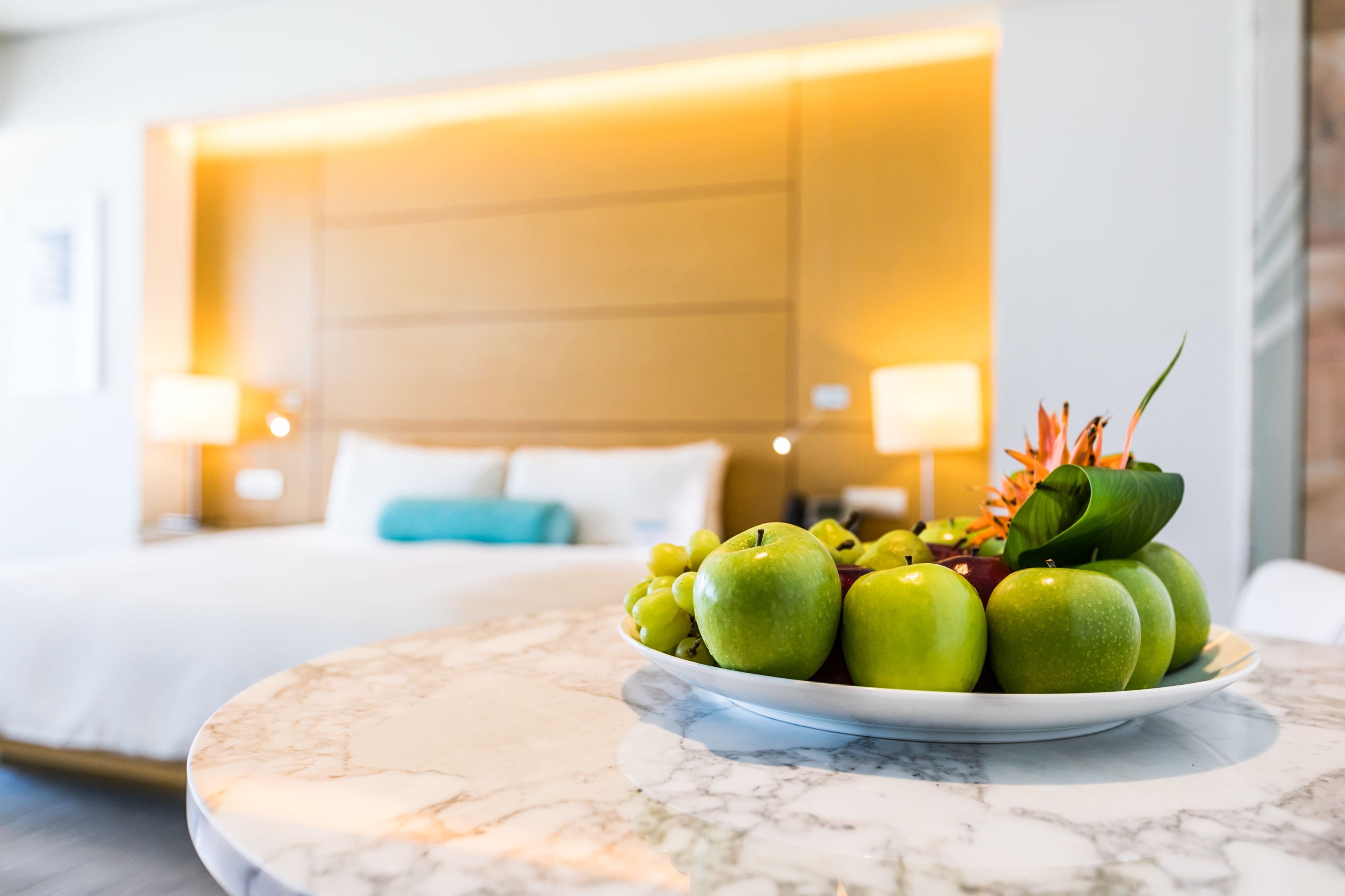 Uma mesa de mármore com um prato de maçãs verdes e uma folha decorativa. Ao fundo, vê-se uma cama com roupa de cama branca e uma cabeceira de madeira.