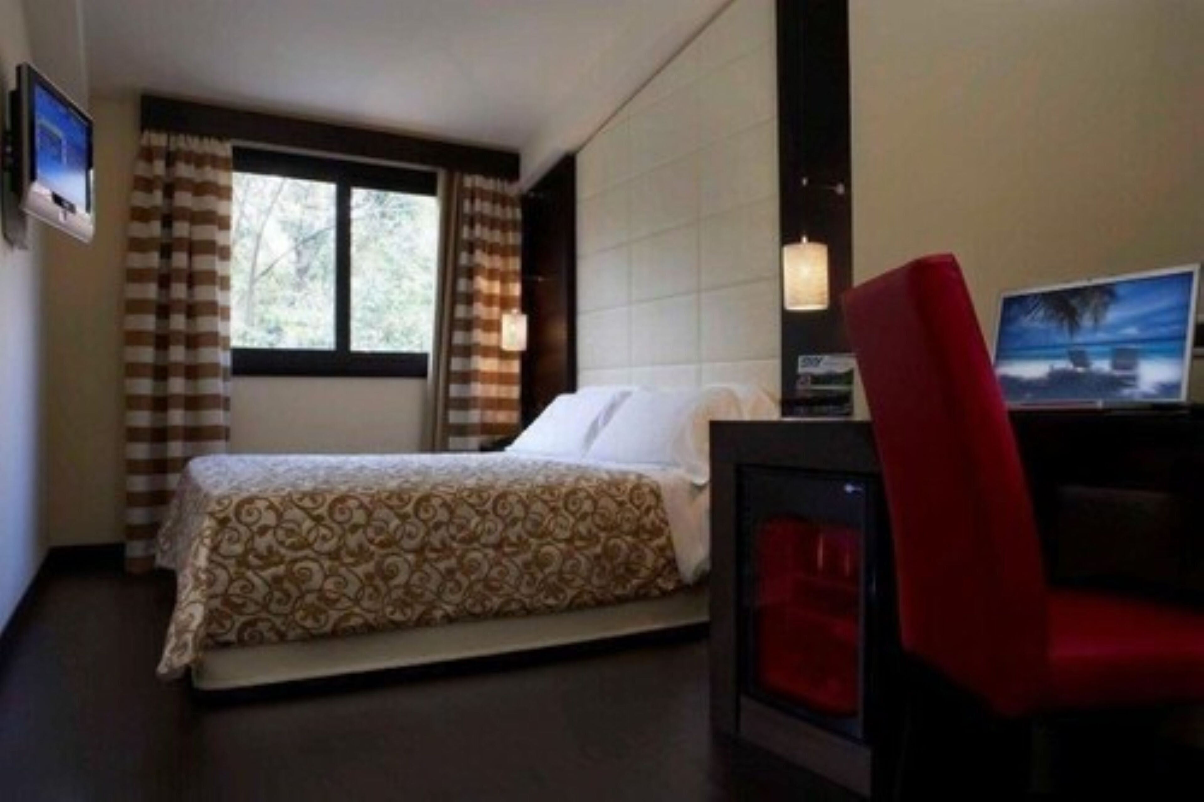 Quarto de hotel com uma cama, uma secretária com cadeira, janela com cortinas e televisão fixada na parede.