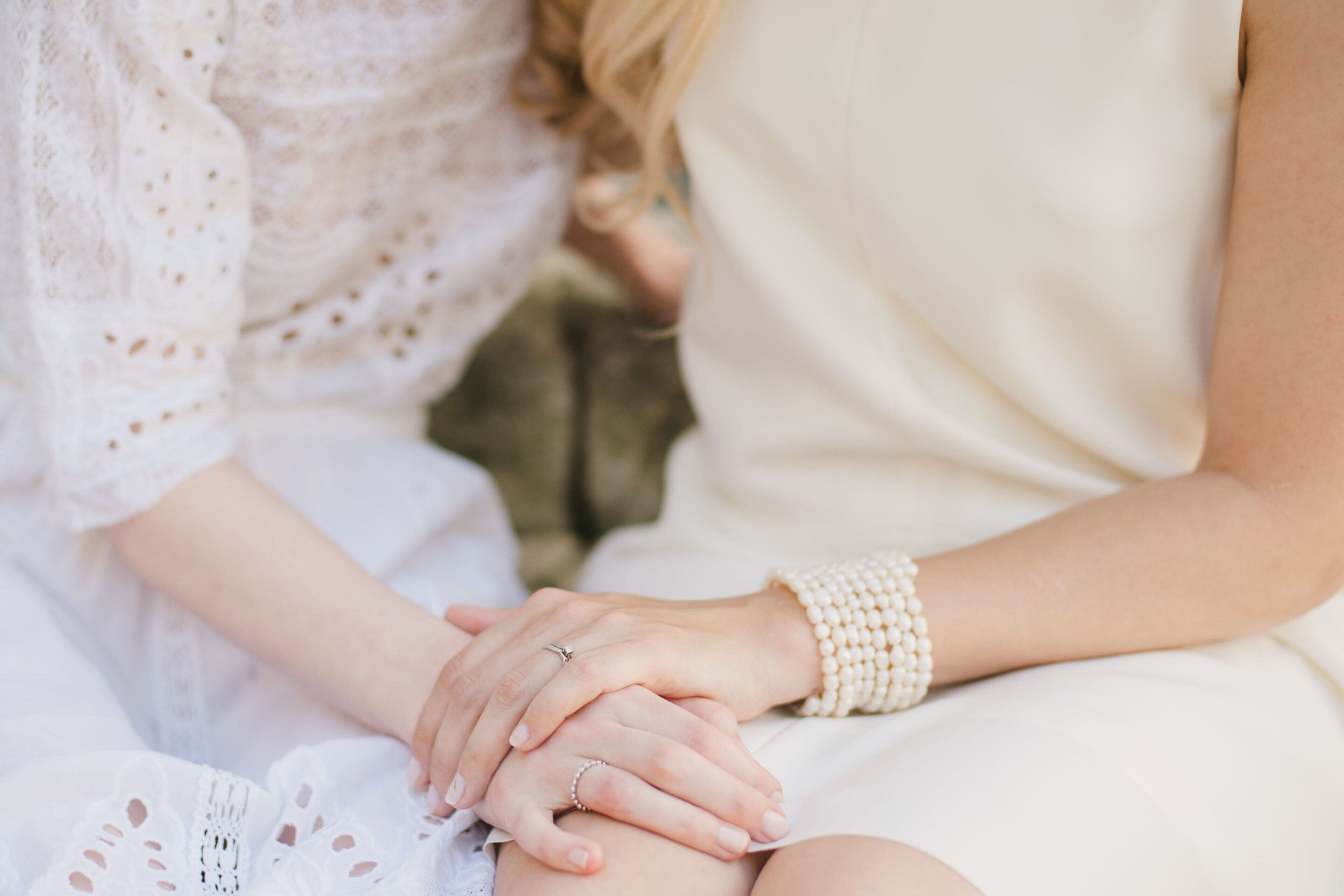 Duas pessoas sentadas perto uma da outra, uma vestindo um vestido branco de renda e a outra, um vestido branco, com as mãos unidas delicadamente.
