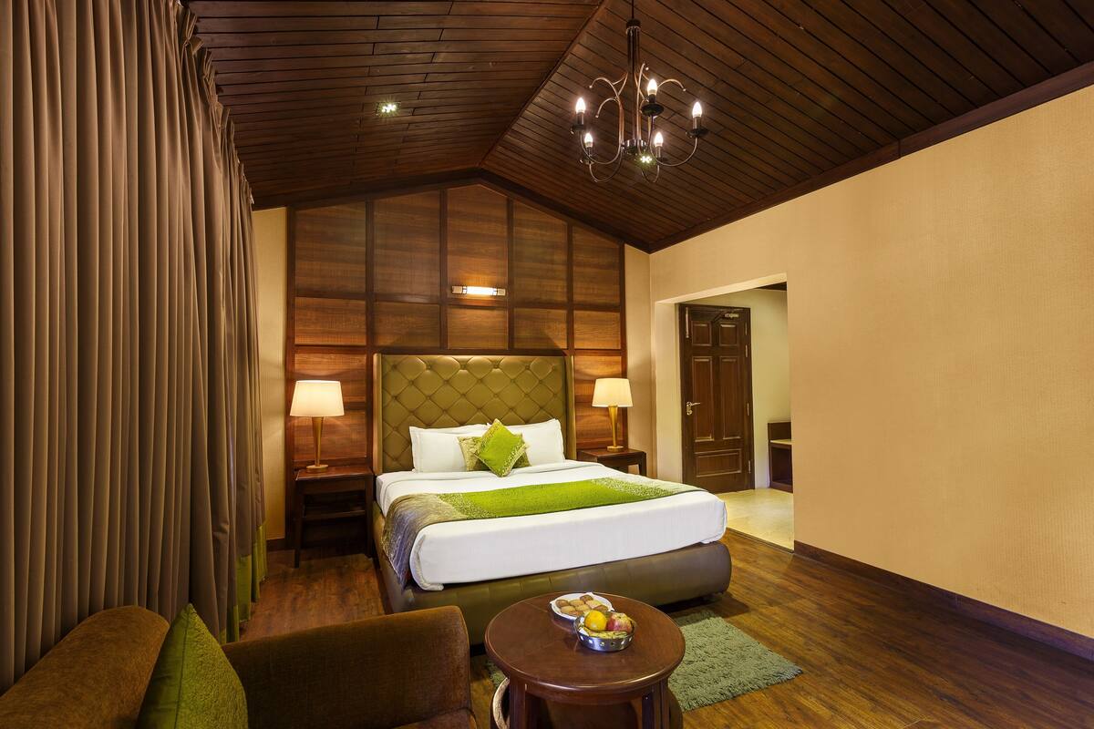 Quarto de hotel com uma cama grande, teto de madeira e um candelabro. Há uma pequena mesa com fruta e um almofado verde.