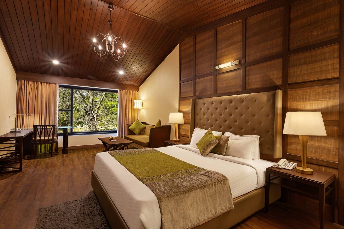 Quarto com uma cama grande, uma escrivaninha e uma cadeira. Há uma janela com cortinas, um lustre e uma mesa de cabeceira com abajur.