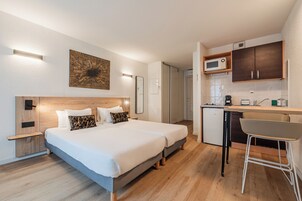 Quarto de hotel com cama, mesas de cabeceira, escrivaninha, cadeira, micro-ondas e pequena cozinha.