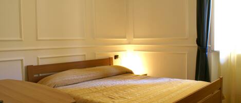 Uma cama de solteiro com cabeceira de madeira e mesa de cabeceira, um abajur na parede e uma janela com cortinas.