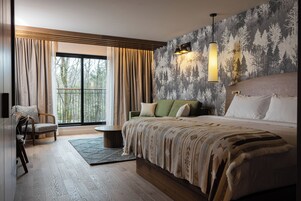 Um quarto moderno com uma cama grande, um sofá verde, piso de madeira e vista para árvores pela janela.