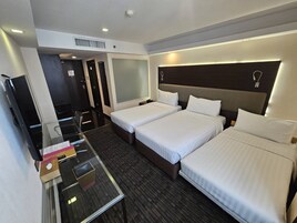 Quarto de hotel com duas camas, uma mesa com computador, uma televisão e um painel de vidro.