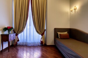 Quarto com piso de madeira, um sofá com almofada, uma mesinha com um vaso e uma janela com cortinas.
