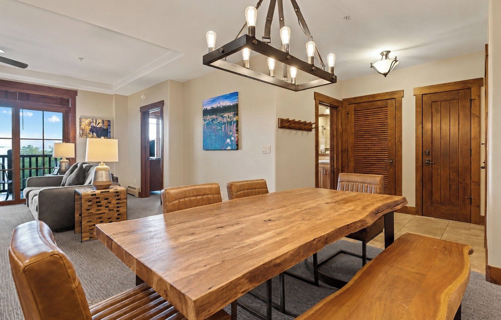 Uma ampla área de jantar com uma grande mesa de madeira e bancos, um candelabro com várias lâmpadas, e vista para o exterior por uma grande janela.