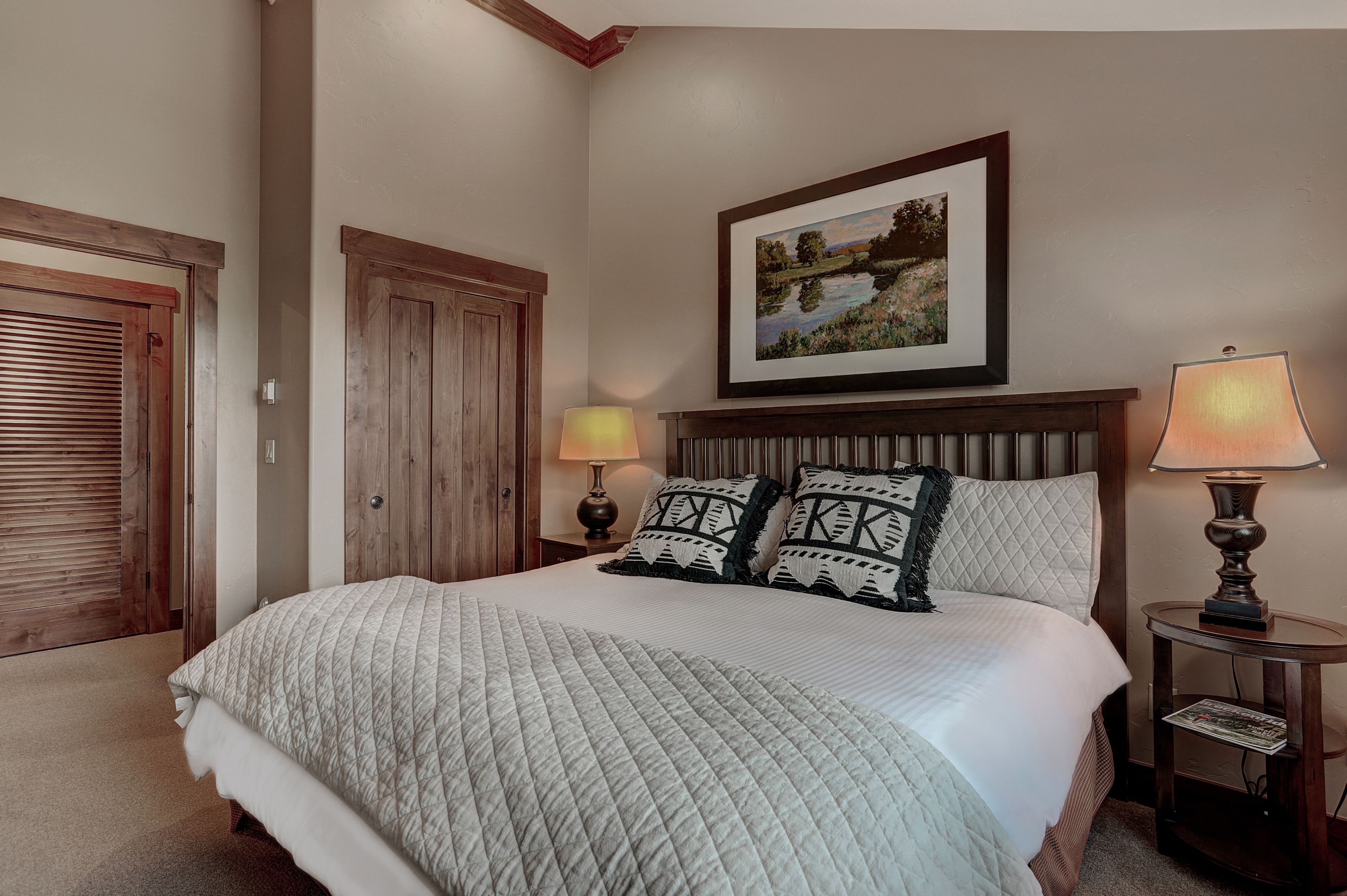 Cama bem arrumada com edredom acolchoado, duas luminárias de cabeceira, um quadro de paisagem acima da cama e uma porta de madeira à esquerda.