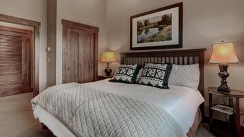 Cama bem arrumada com edredom acolchoado, duas luminárias de cabeceira, um quadro de paisagem acima da cama e uma porta de madeira à esquerda.