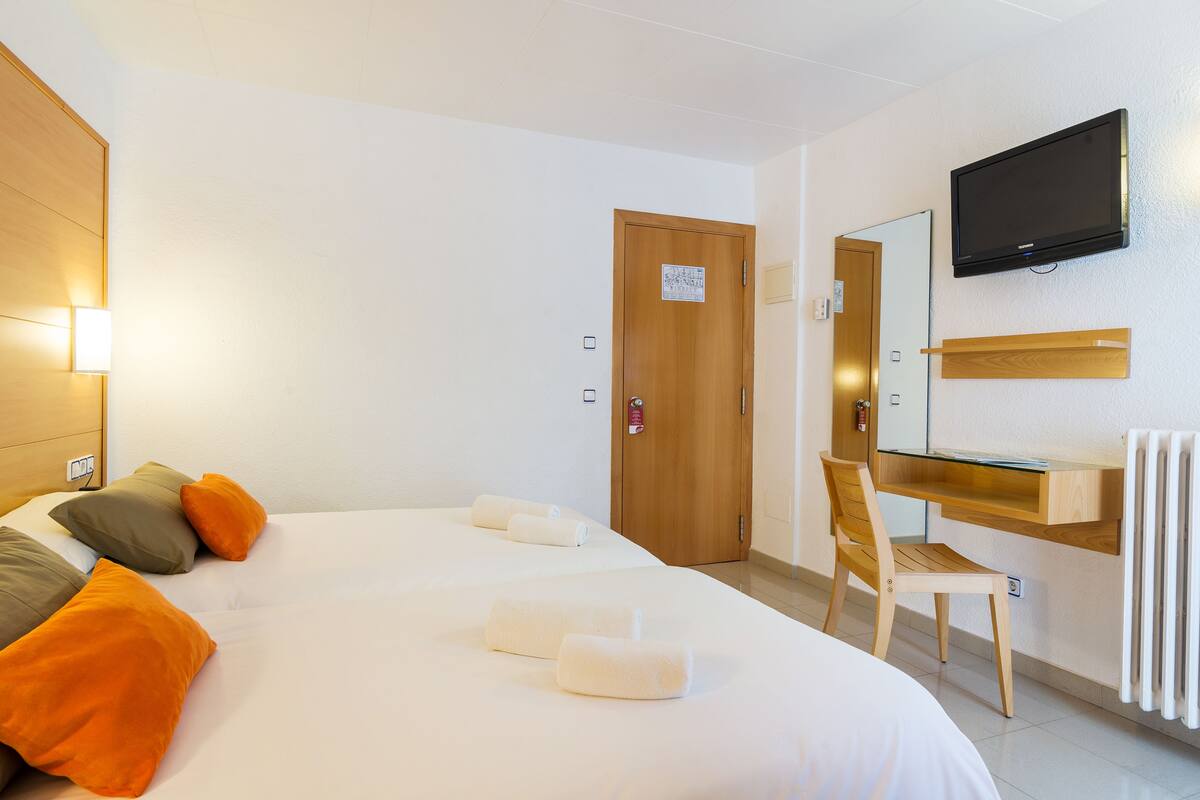 Quarto de hotel com cama, escrivaninha, cadeira, televisão e espelho.