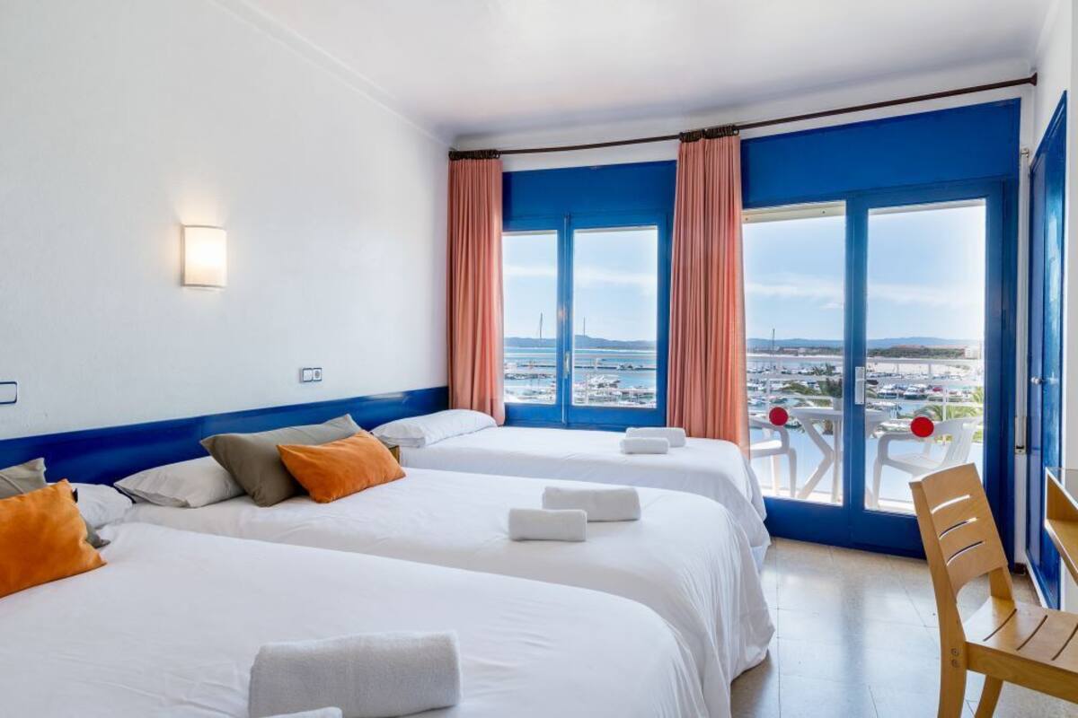 Quarto de hotel com duas camas, uma escrivaninha e uma cadeira. Há uma janela ampla com vista para um porto e edifícios.