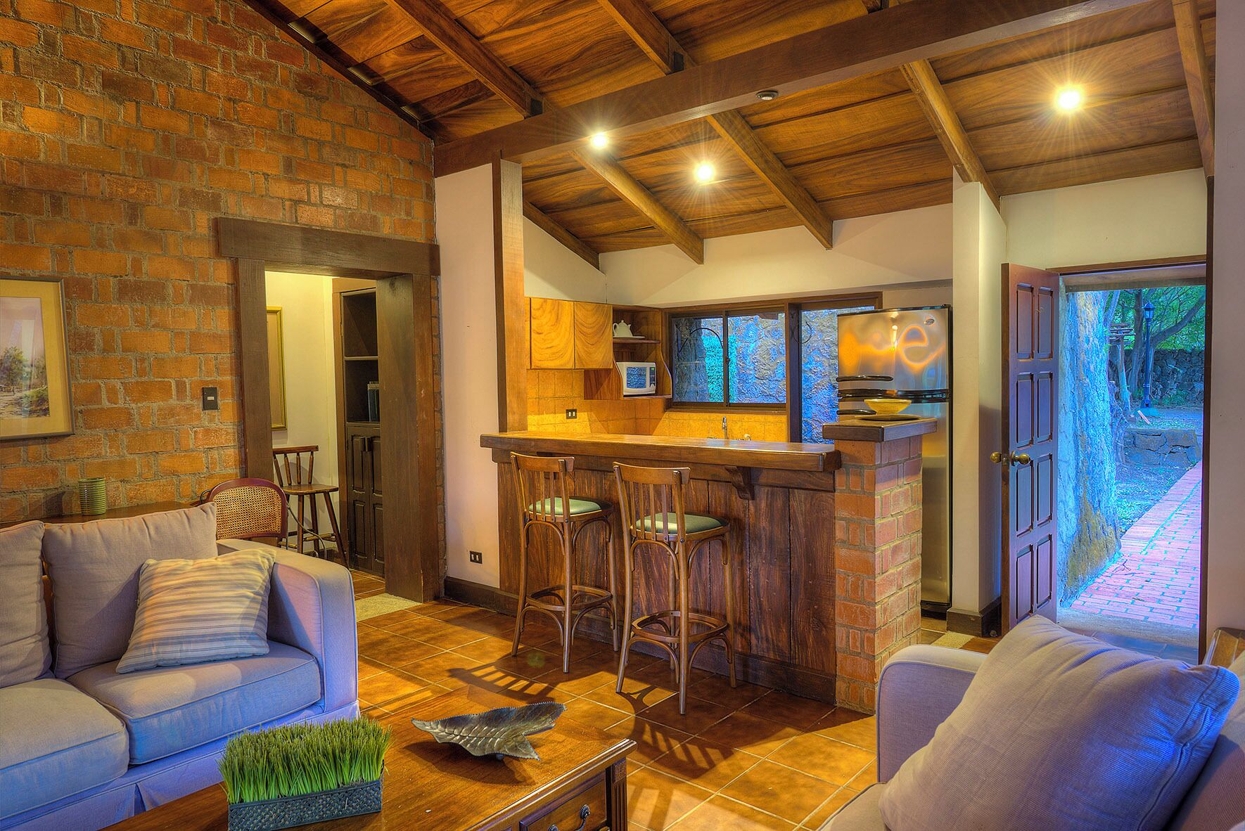 Uma sala de estar aconchegante com sofá e poltronas, uma cozinha com bancada e vista para um jardim através de uma porta aberta.