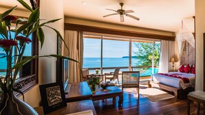 Quarto com cama, escrivaninha, cadeira, mesa, planta e vista para o oceano.