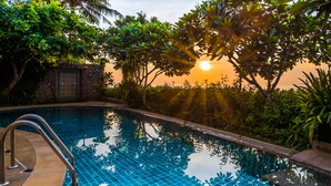 Uma piscina com muro de pedra e corrimão de metal, cercada por vegetação exuberante e um pôr do sol ao fundo.
