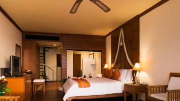 Quarto de hotel com cama, escrivaninha com televisão, cadeira, mesa de cabeceira com abajur e ventilador de teto.