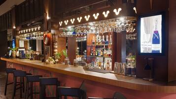 Uma área de bar com balcão de madeira, banquetas e diversas garrafas e copos.