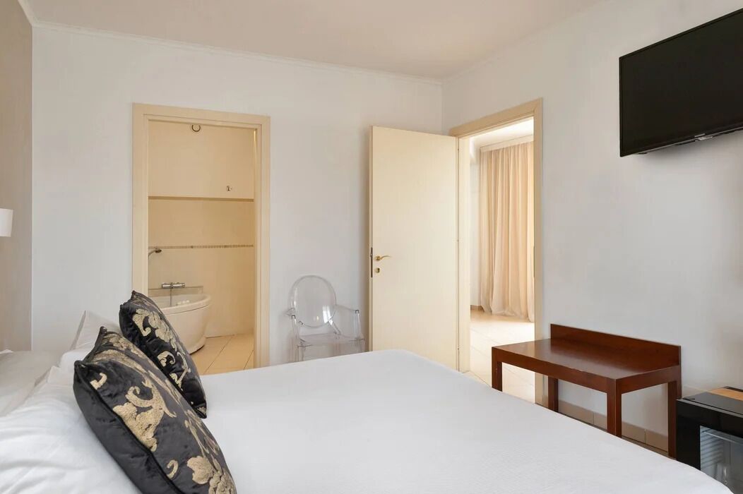 Quarto de hotel com uma cama, uma mesa de madeira, uma televisão fixada na parede, um banheiro com vaso sanitário e pia, e uma cadeira.