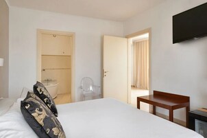 Quarto de hotel com uma cama, uma mesa de madeira, uma televisão fixada na parede, um banheiro com vaso sanitário e pia, e uma cadeira.