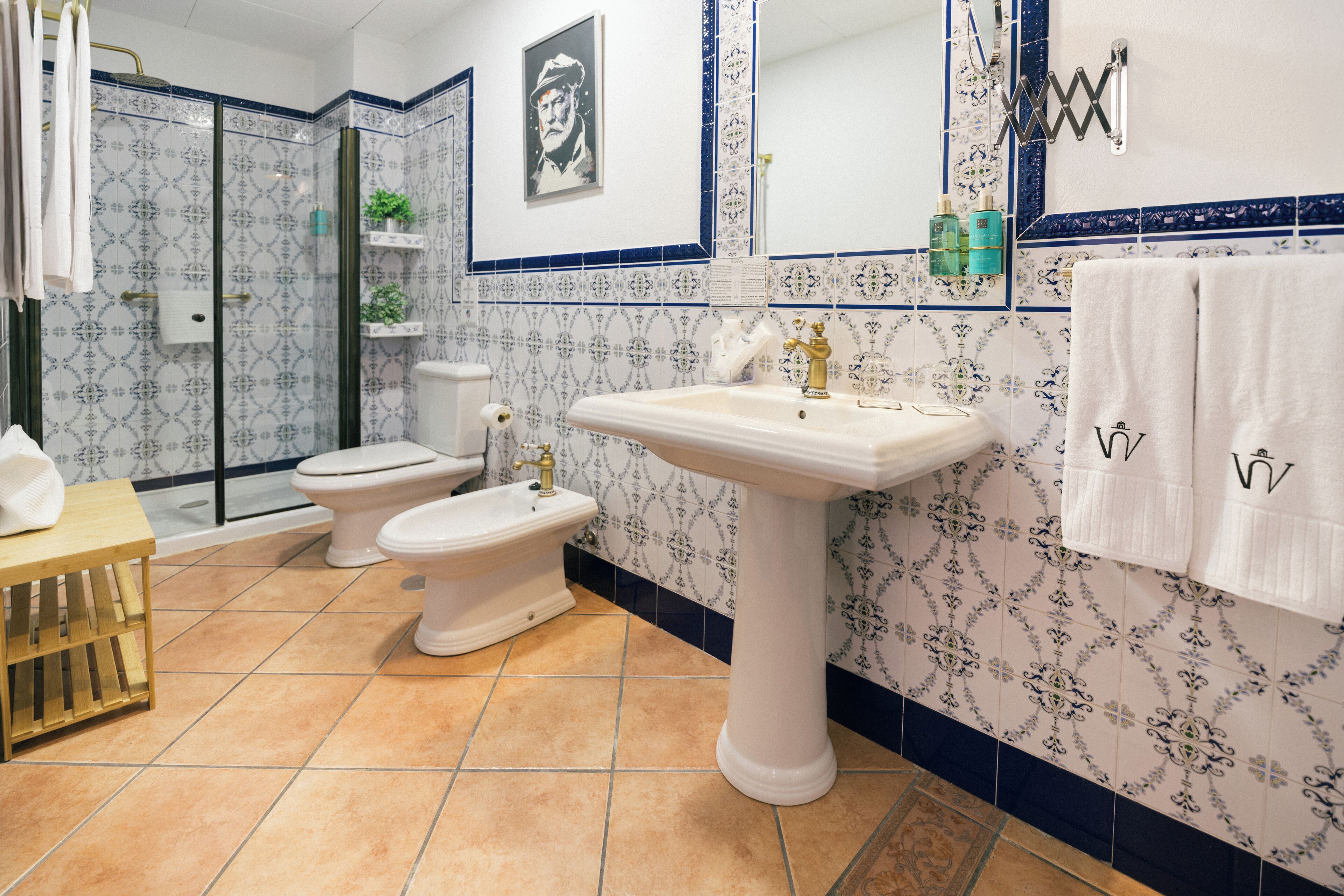 Uma casa de banho com lavatório pedestal branco, duas sanitas, zona de duche e chão em mosaico.
