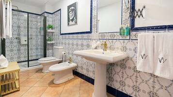 Uma casa de banho com lavatório pedestal branco, duas sanitas, zona de duche e chão em mosaico.
