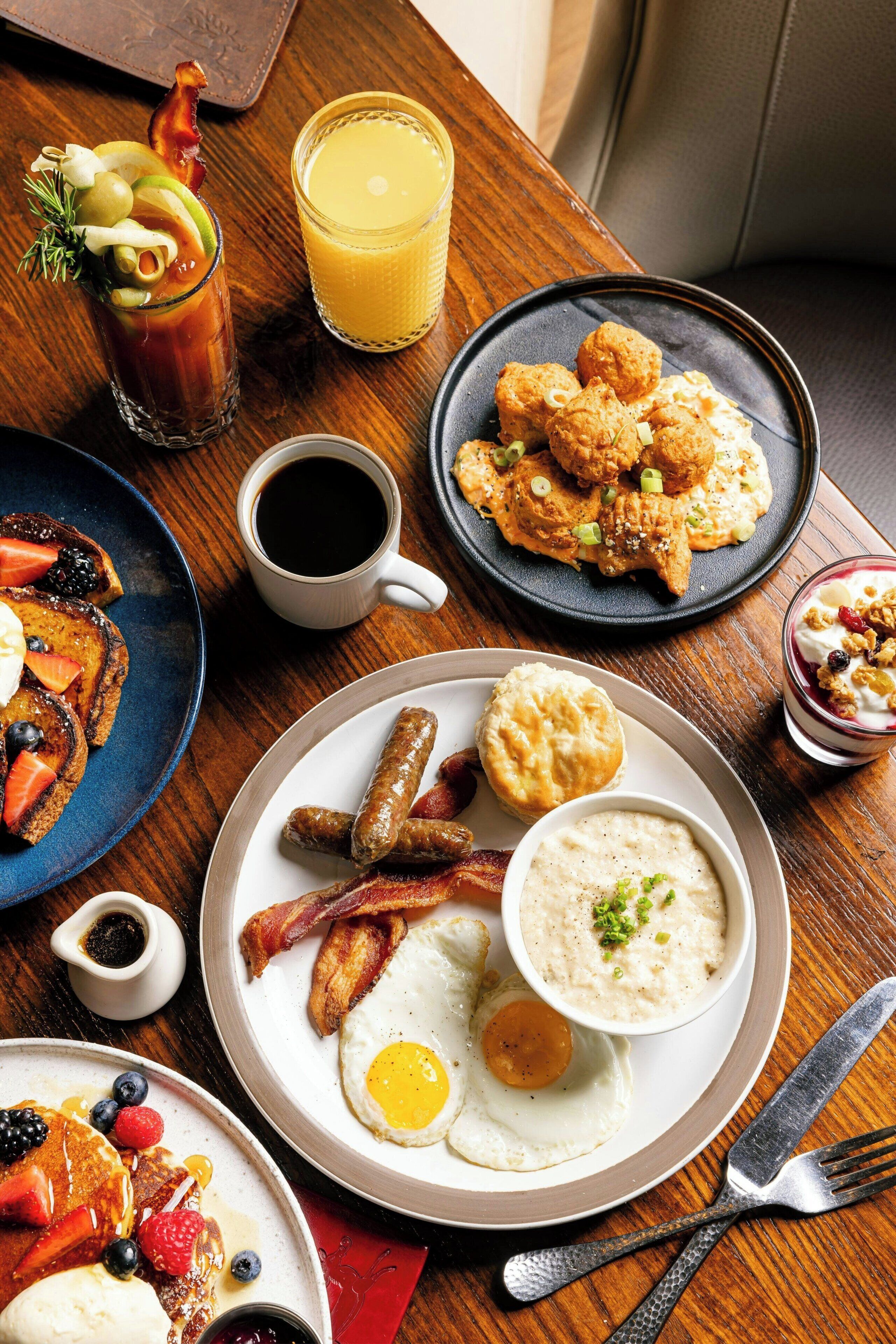 Uma mesa de café da manhã com diversos pratos, incluindo ovos, linguiça, panquecas e um drink Bloody Mary.