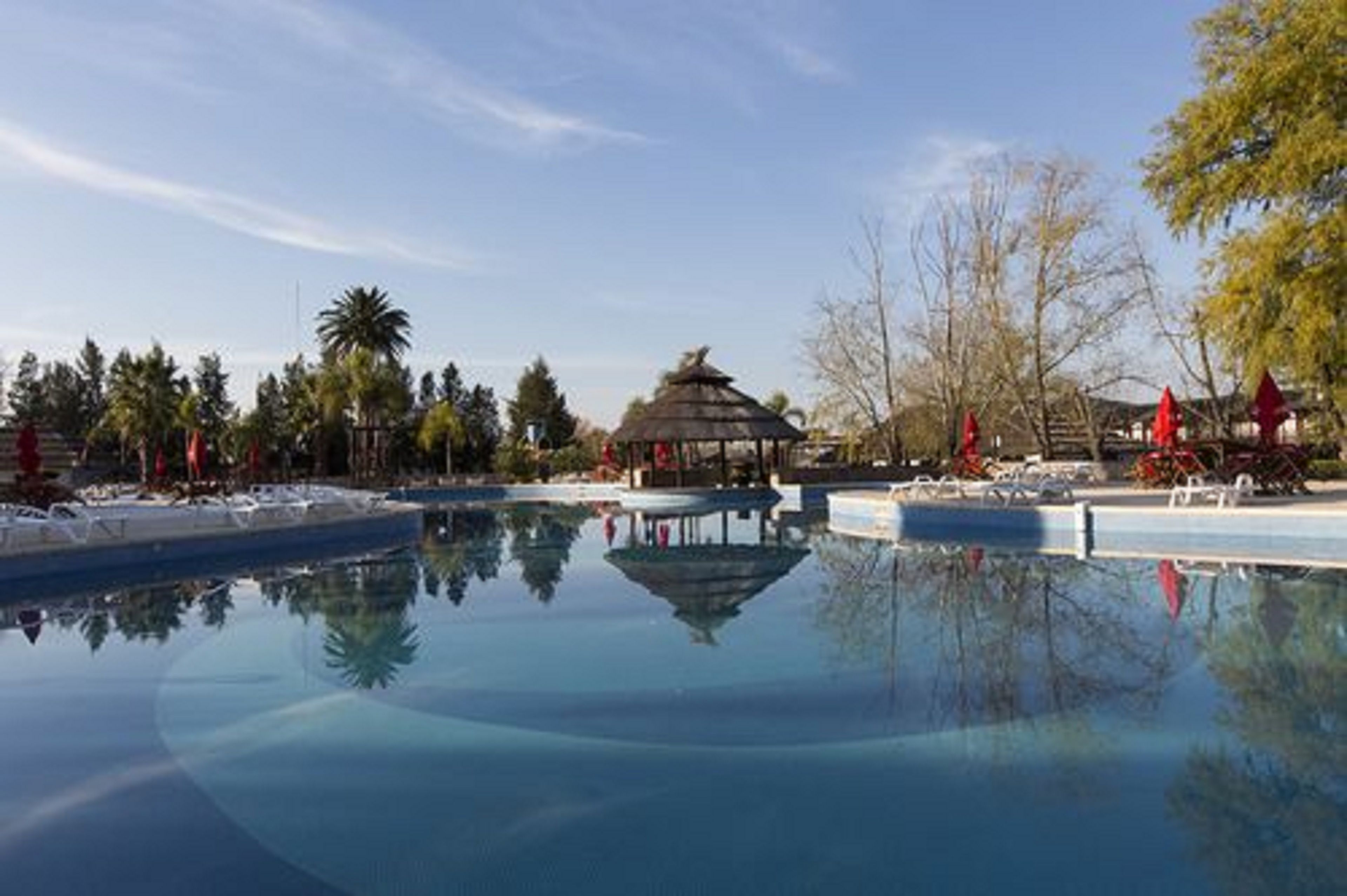 Piscina cristalina com um gazebo no centro, rodeada por palmeiras e espreguiçadeiras.