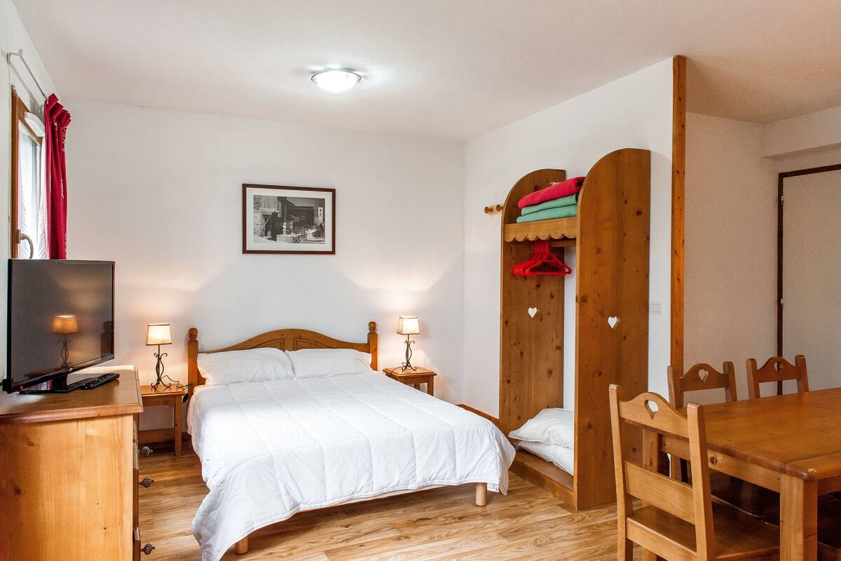 Quarto com cama, mesas de cabeceira, televisão, guarda-roupa de madeira e uma mesa de jantar com cadeiras.