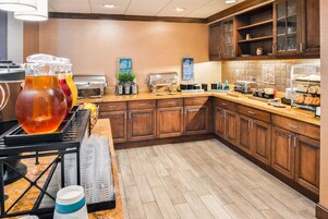 Uma cozinha de hotel com buffet, incluindo um espaço para bebidas com jarras e copos, uma torradeira e uma vitrine com pães e doces.