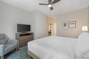 Quarto de hotel com uma cama, uma televisão sobre uma cômoda de madeira, um ventilador de teto e duas fotos emolduradas na parede.