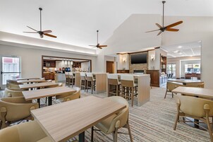 Um espaço de refeições de hotel com várias mesas e cadeiras, ventiladores de teto, área de cozinha e uma televisão na parede.