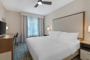 Quarto de hotel com uma cama, uma escrivaninha com computador, um ventilador de teto, uma janela com cortinas e uma mesa de cabeceira com abajur.