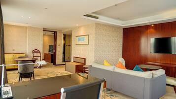 Quarto de hotel com área de estar, mesa de jantar, sofá com almofadas, televisão na parede e escrivaninha com cadeira.
