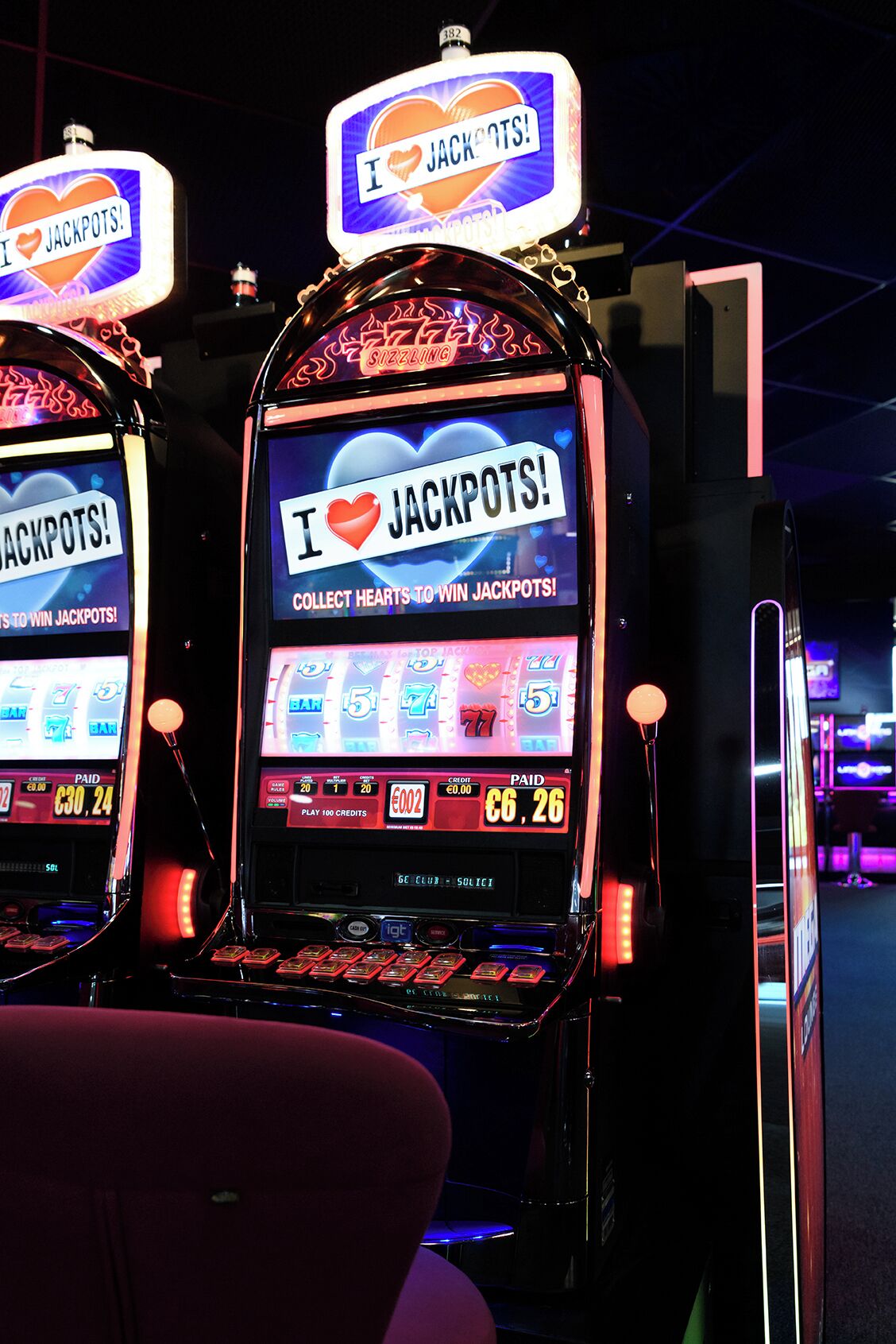 Duas máquinas caça-níqueis com letreiros iluminados que dizem "I ♥ JACKPOTS!" e "Colecione corações para ganhar jackpots!"